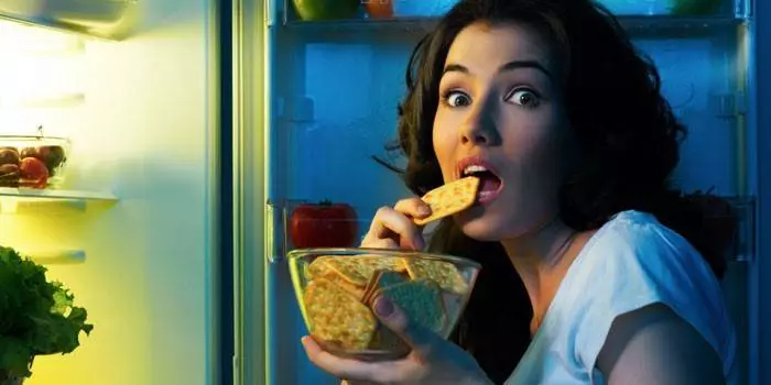 Дівчина перед відкритим холодильником їсть крекери
